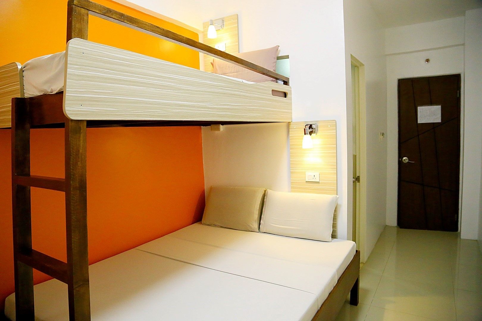 Uma cama de solteiro com beliche inferior e sofá-cama, uma luminária fixada na parede e uma porta em um quarto com paredes laranja.