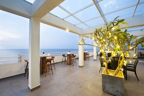 Amplo espaço interno com vista para o mar, com mesas e cadeiras dispostas para refeições, e uma grande planta em vaso com iluminação.