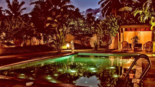 Piscina noturna com uma estátua, cercada por palmeiras e um edifício com área de estar externa.