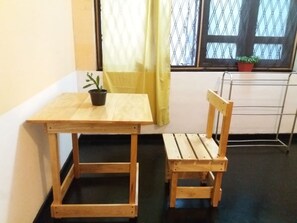 Uma pequena mesa de madeira com um vaso de planta e uma cadeira em um quarto com janela e um suporte de metal.