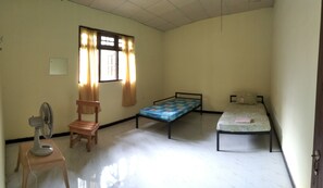 Quarto com duas camas de solteiro, uma cadeira de madeira, um ventilador e uma janela com cortinas.