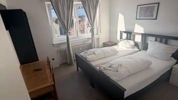 Quarto de hotel com uma cama, uma escrivaninha, uma cadeira, uma janela com cortinas, um radiador e um quadro na parede.