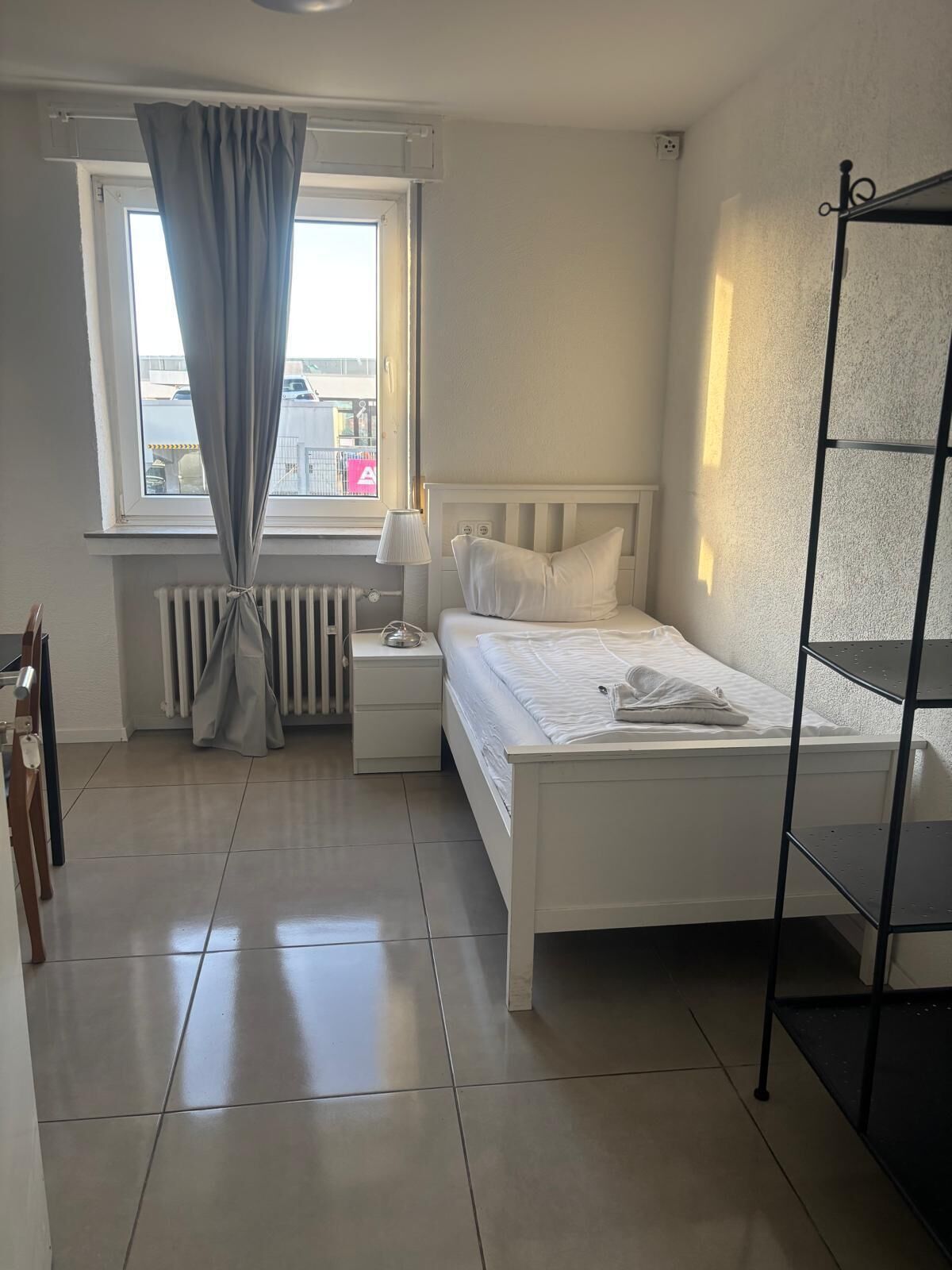 Quarto com cama, mesa de cabeceira, radiador, janela com cortinas e estante de metal.