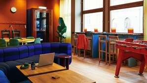 Uma área de estar moderna com sofá azul, mesa de centro de madeira, um laptop, mesa de pebolim e um painel de mensagens.