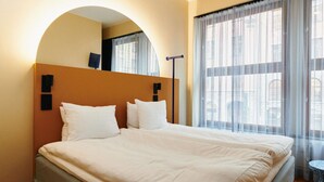 Quarto de hotel com uma cama grande, cabeceira, mesa de cabeceira e janela com cortinas leves.