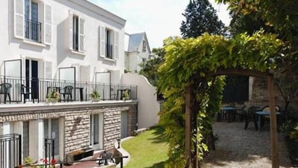 Um edifício branco com fachada de pedra, uma varanda com grades de metal e um jardim com mesas e cadeiras.