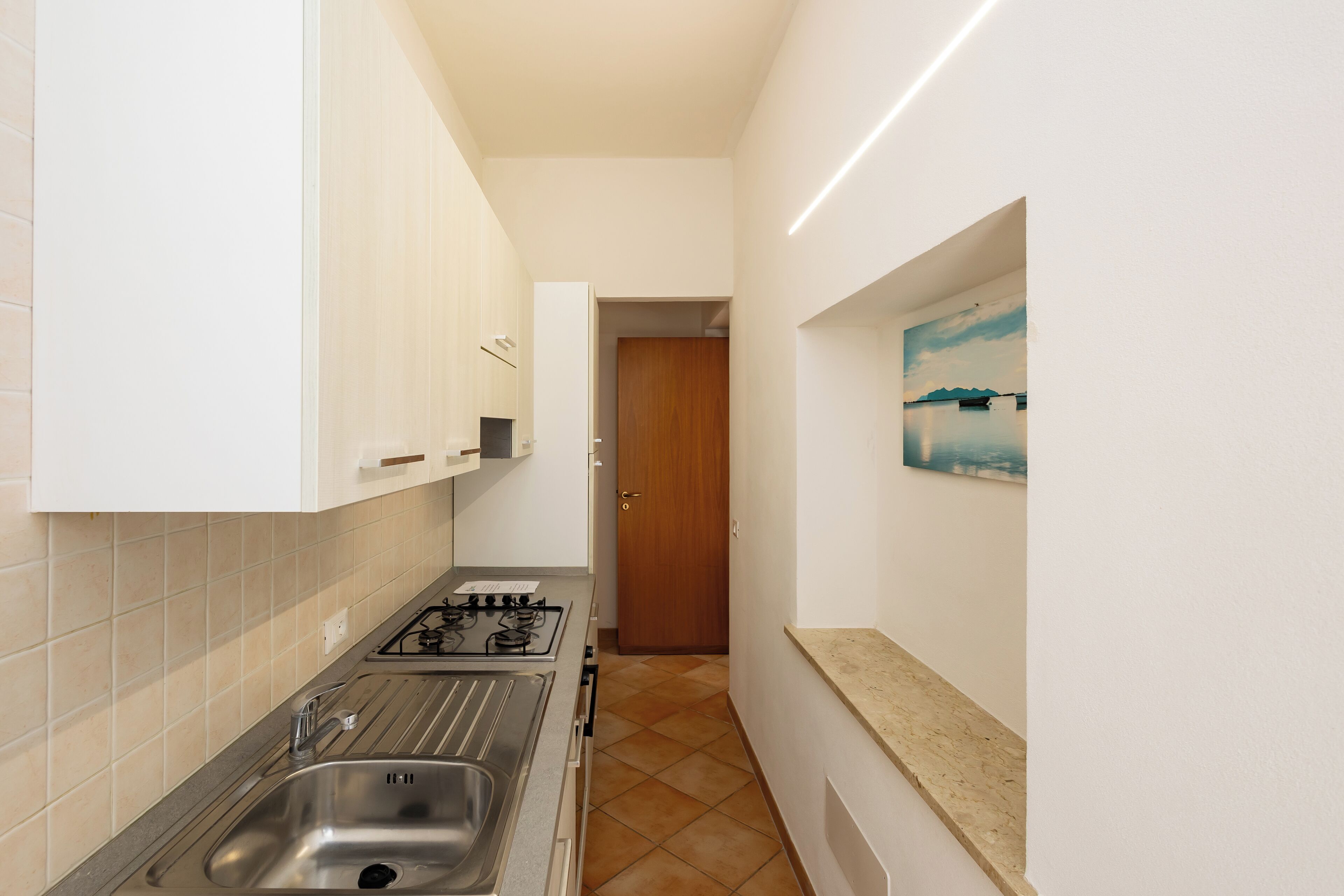 Cozinha pequena com armários brancos, pia de aço inoxidável e painel de azulejos.