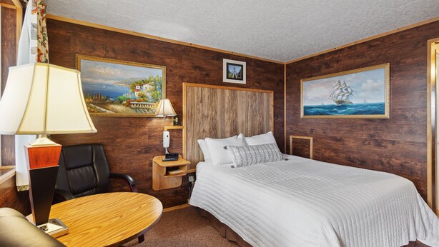 Quarto de hotel com cabeceira de madeira, cama com roupa de cama branca, mesa de madeira, uma cadeira e um abajur.