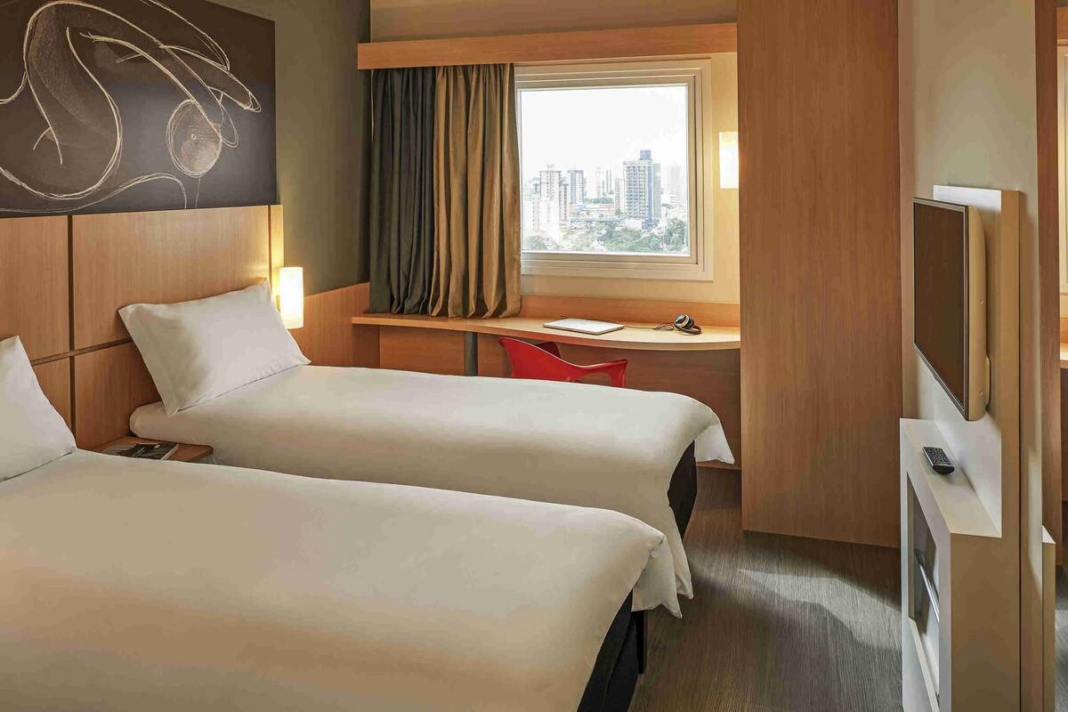 Quarto de hotel com duas camas, uma escrivaninha, uma televisão e uma janela com vista para a cidade.