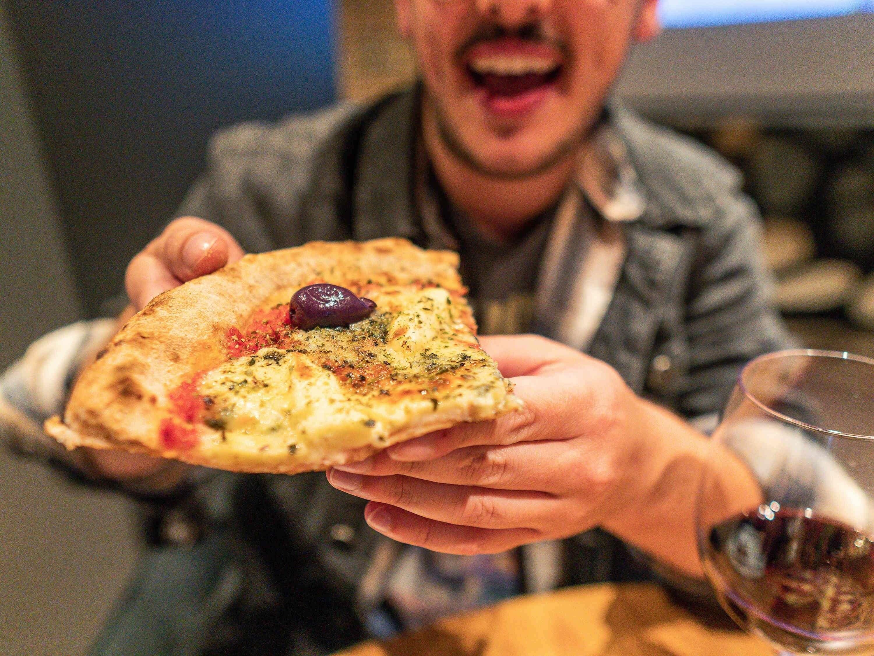 Uma pessoa segurando uma fatia de pizza com cobertura vermelha e uma azeitona preta.
