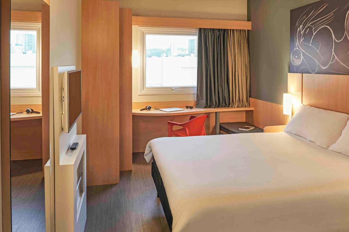 Quarto de hotel com cama, escrivaninha, cadeira e janela com cortinas.