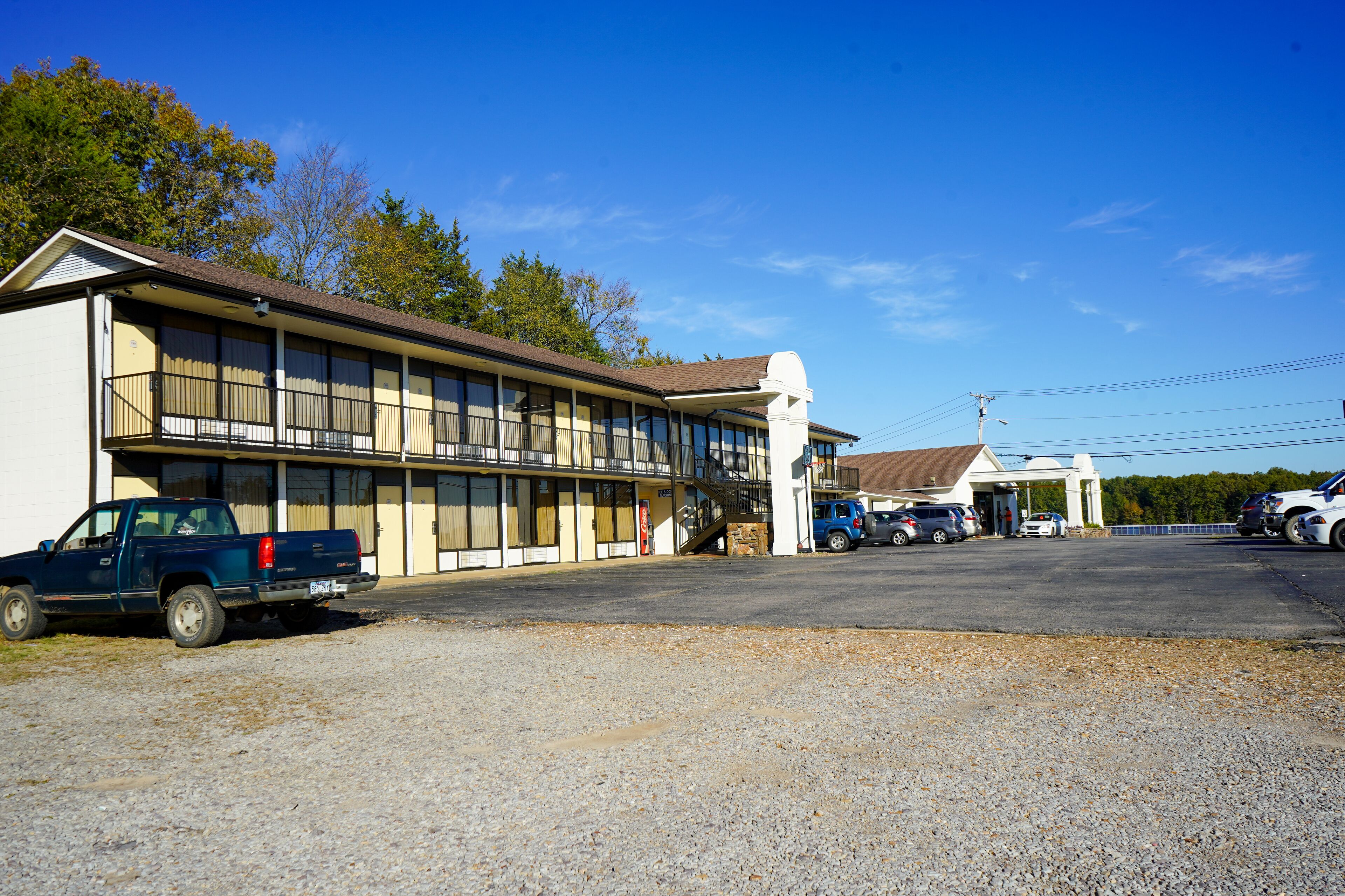 Um motel com um prédio de dois andares, área de estacionamento coberta e entrada com calçamento de brita.