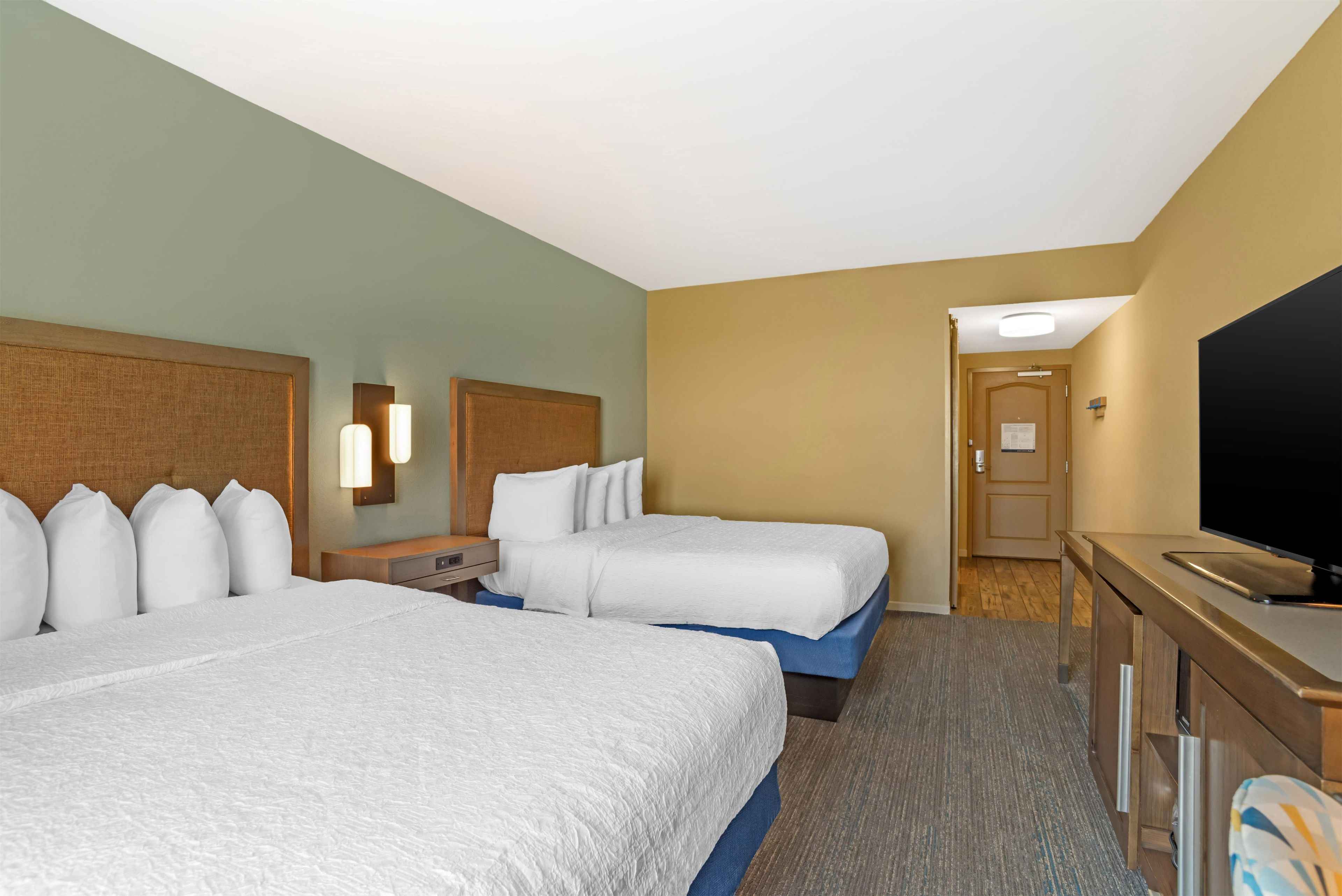 Quarto de hotel com duas camas, uma escrivaninha e uma televisão de tela plana, e uma porta que dá para um corredor.