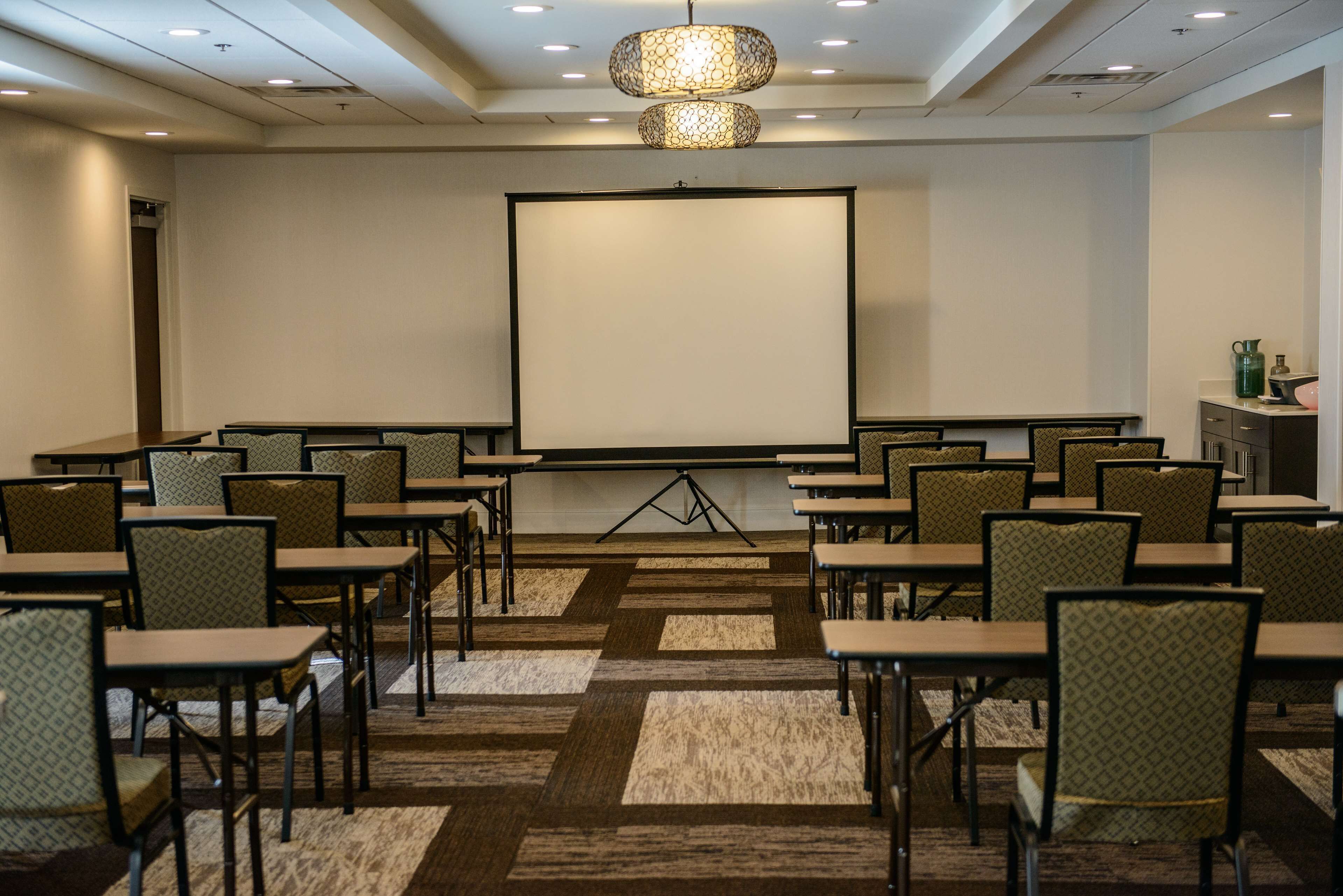 Uma sala de conferências com uma tela grande, várias mesas e cadeiras dispostas para uma reunião.