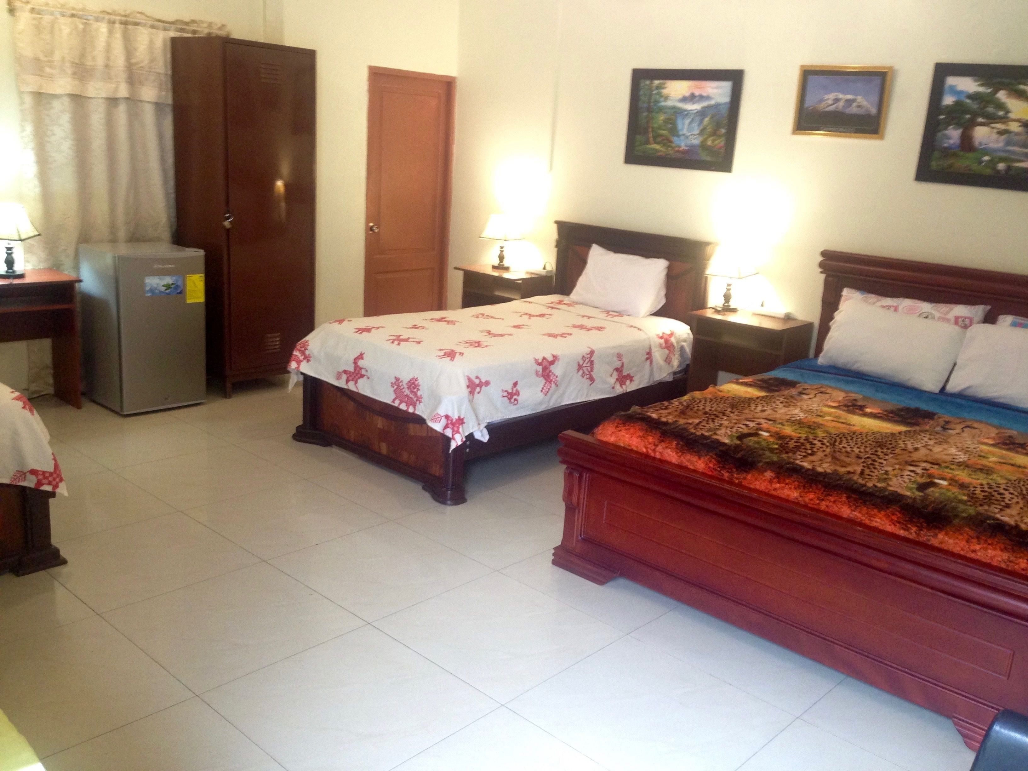 Quarto de hotel com duas camas, um frigorífico, um guarda-roupa e uma secretária com uma lâmpada.