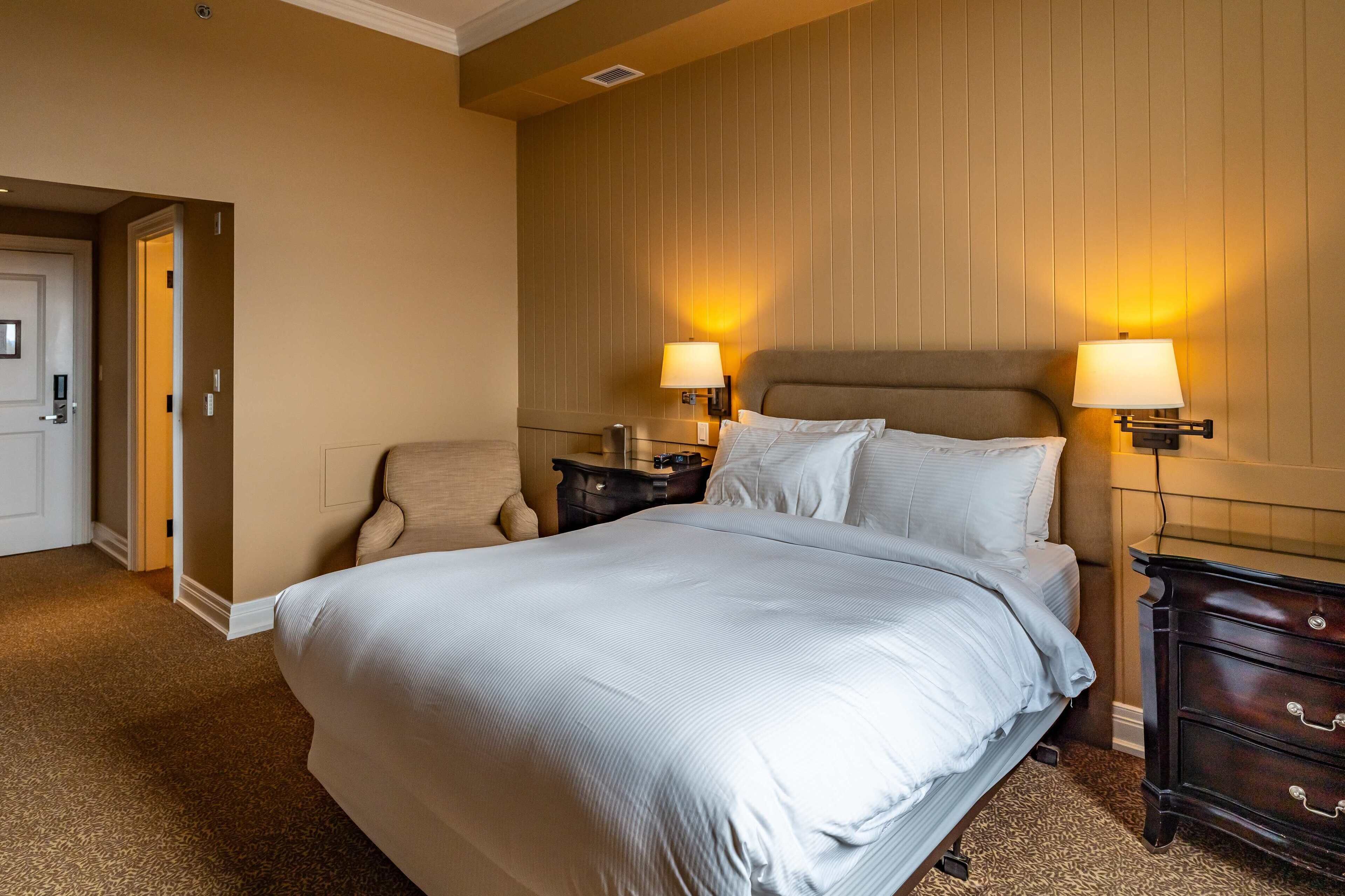 Quarto de hotel com uma cama grande, duas luminárias de cabeceira, cômoda de madeira escura e uma cadeira.