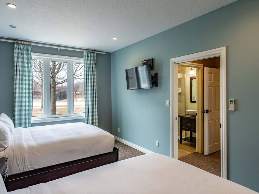 Quarto com duas camas, janela com cortinas, televisão na parede e porta para um banheiro.