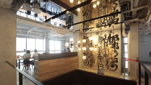 Um interior moderno com um balcão de recepção de madeira, uma grande parede decorativa e uma escadaria com guarda-corpo de metal.