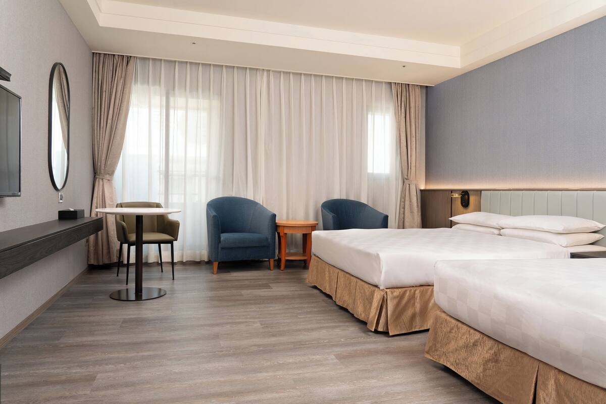 Quarto de hotel com uma cama, duas cadeiras, uma mesinha, um espelho e uma televisão.