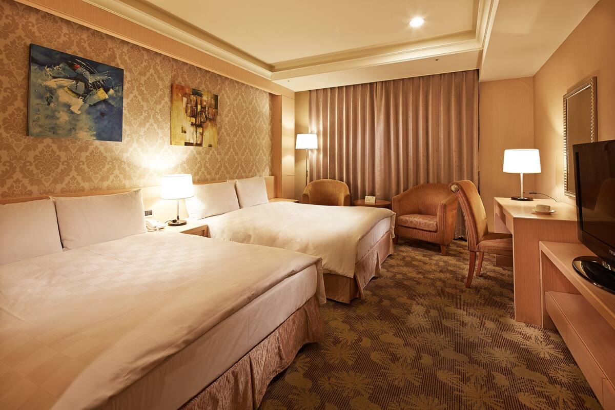 Quarto de hotel com duas camas, uma mesa de trabalho com televisão, uma cadeira e obras de arte nas paredes.