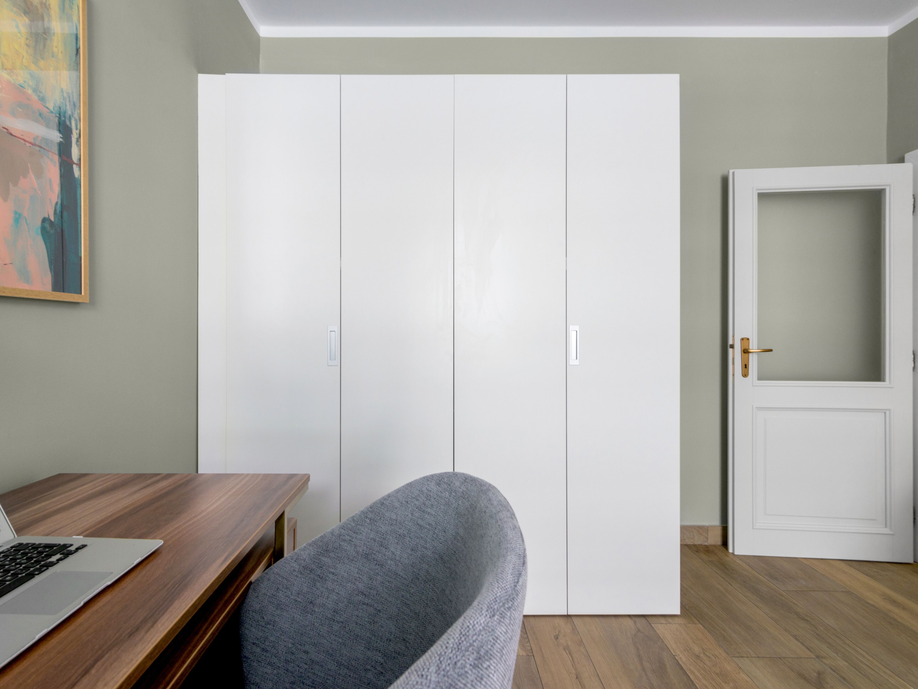 Um quarto com escrivaninha de madeira, uma cadeira cinzenta, um guarda-roupa branco e uma porta com painel de vidro.