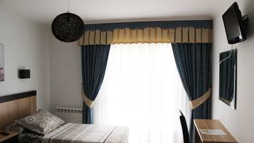 Quarto de hotel com cama, escrivaninha, cadeira, televisão e janela com cortinas.