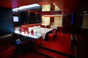 Quarto de hotel moderno com divisória de vidro, uma cama, uma escrivaninha e uma poltrona estofada vermelha.