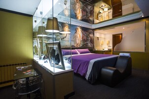 Quarto de hotel com divisória de vidro, cama com roupa de cama roxa, escrivaninha com luminária e banheiro com espelho e porta-toalhas.