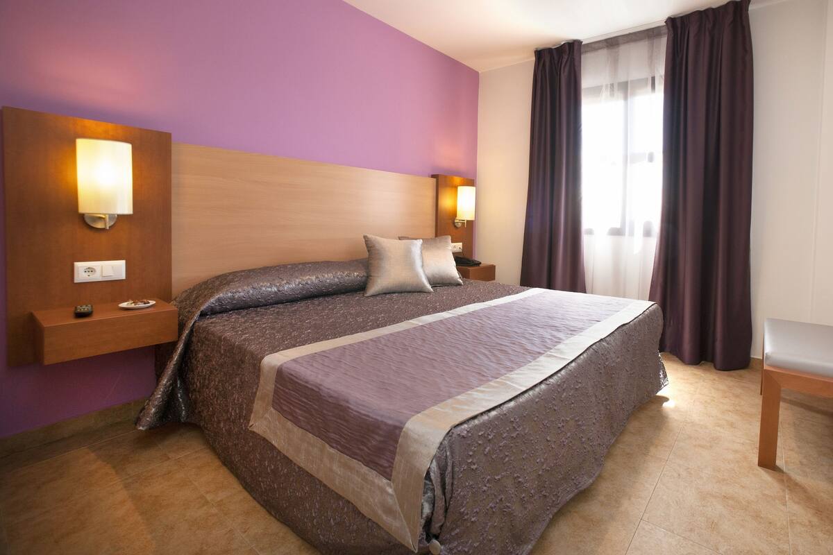 Quarto de hotel com uma cama grande, mesas de cabeceira, uma cadeira e uma TV fixada na parede.