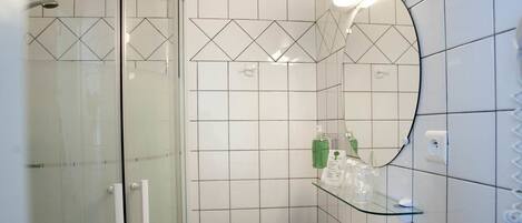 Casa de banho moderna com cabine de duche em vidro, lavatório branco e estante de vidro com artigos de higiene pessoal.
