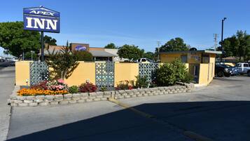 Entrada de um hotel com a placa do Apex Inn, apresentando uma entrada com portão, canteiros de flores e uma área de estacionamento.