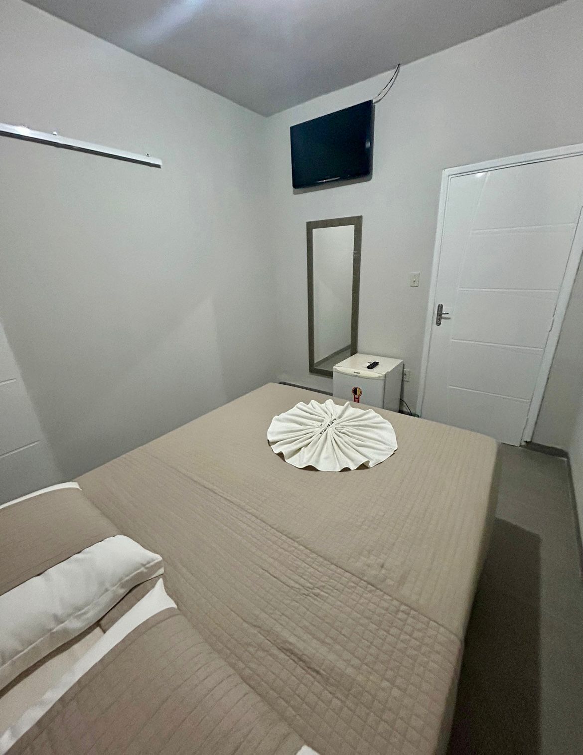 Quarto com cama, mesa de cabeceira, espelho, televisão na parede e uma porta.