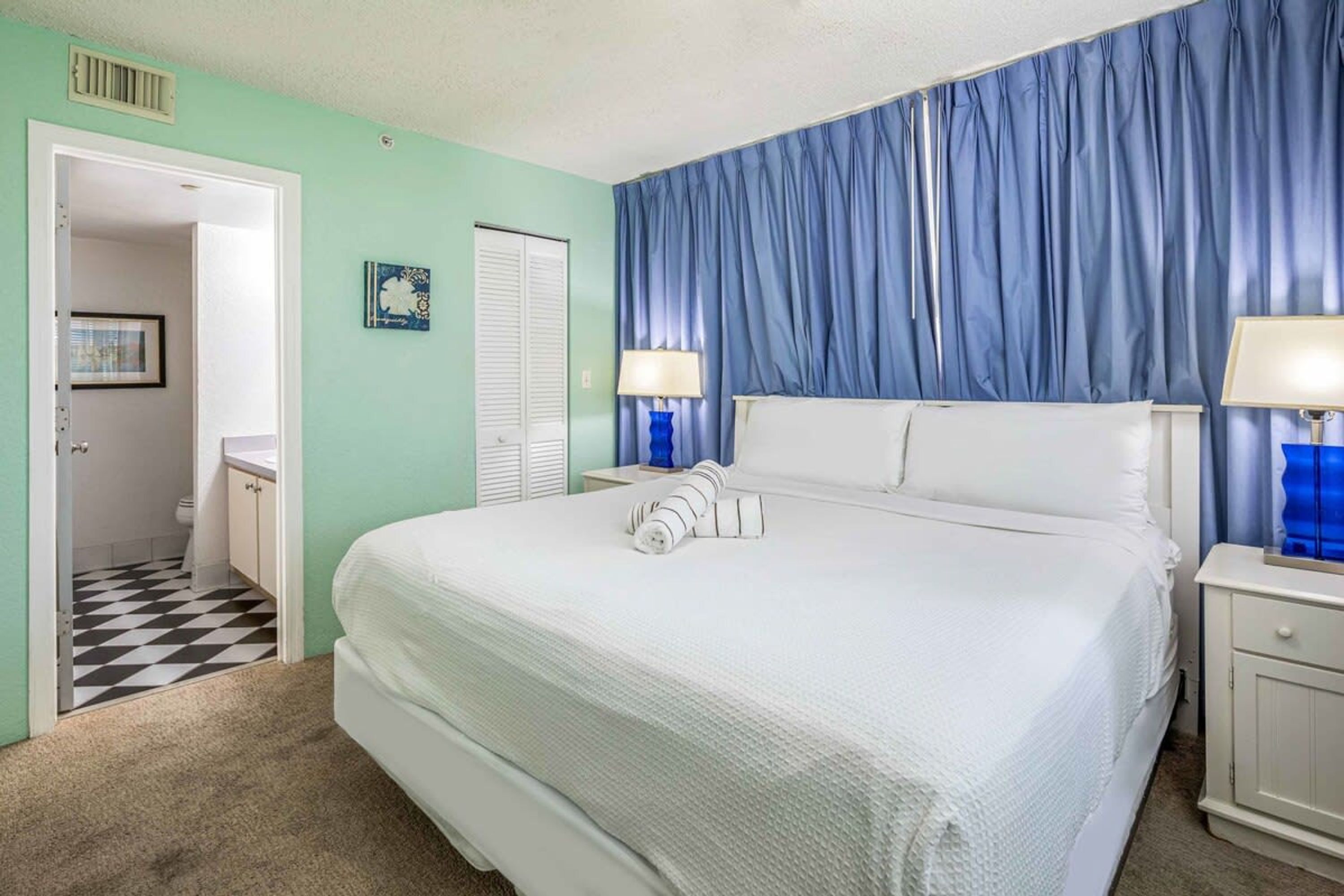 Quarto com uma cama grande, uma mesinha de cabeceira, uma luminária, um banheiro com piso xadrez e uma cortina azul.