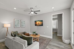 Uma sala de estar moderna com um sofá cinza, uma mesa de centro e um ventilador de teto. Há um quadro na parede e uma porta que dá para outro cômodo.
