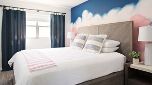 Uma cama bem arrumada com cabeceira de madeira, mesa de cabeceira com luminária e um mural de nuvens na parede.