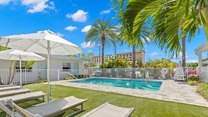 Área de piscina com espreguiçadeiras e guarda-sóis, cercada por palmeiras e com isolamento na piscina.