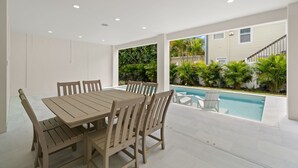 Uma área moderna de refeições ao ar livre com piscina, rodeada por vegetação exuberante e uma escadaria que leva a um segundo andar.