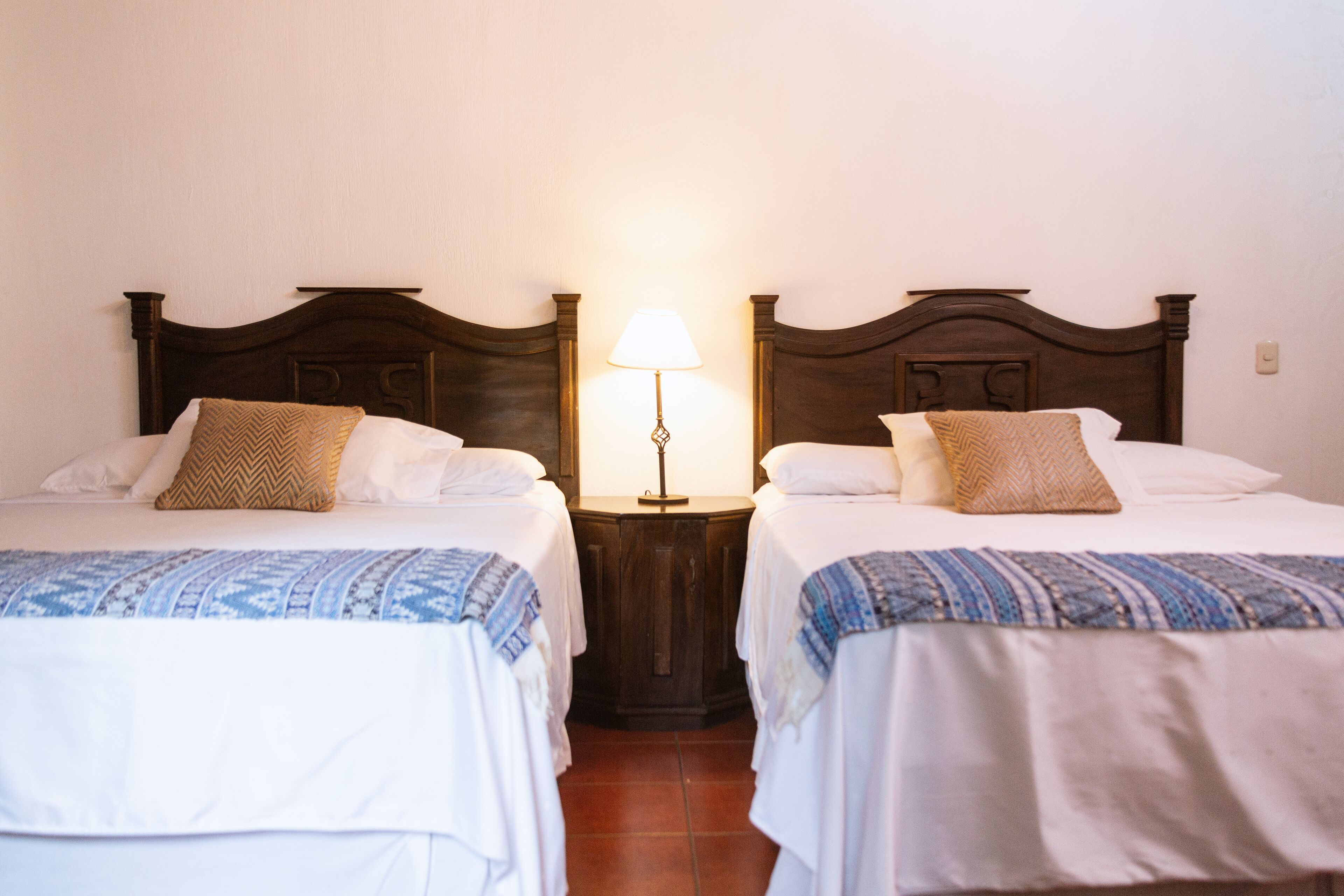 Quarto com duas camas de solteiro, cada uma com um edredom estampado e uma almofada. Um criado-mudo com abajur fica entre as camas.