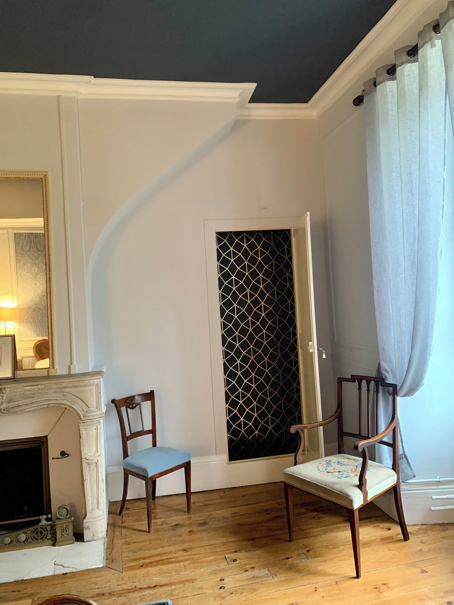 Um quarto com lareira, duas cadeiras de madeira, uma porta com padrão geométrico e uma janela com cortinas.