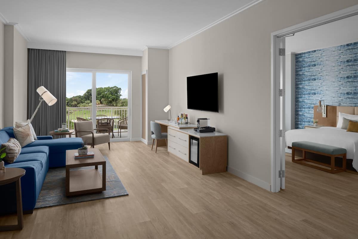 Uma sala de estar moderna com um sofá azul, uma mesa de centro, uma escrivaninha com um computador, uma TV fixada na parede e uma varanda com vista para o verde.