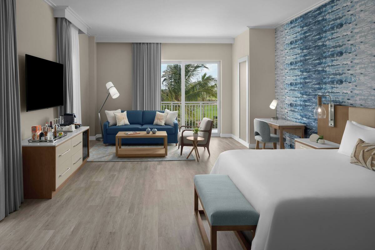 Quarto de hotel moderno com uma cama grande, um sofá, uma mesa de trabalho e uma varanda com vista para palmeiras.