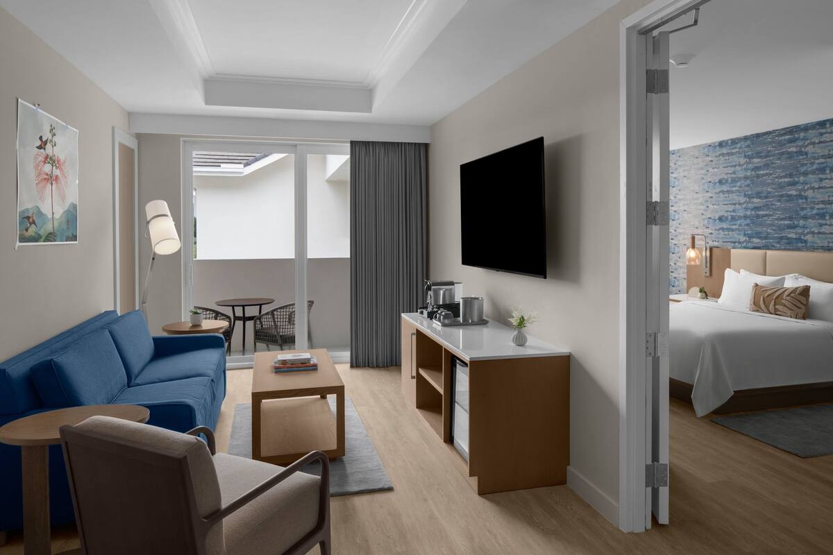 Quarto de hotel moderno com sofá, mesa de centro, escrivaninha, televisão de tela plana, cama e vista para o exterior.