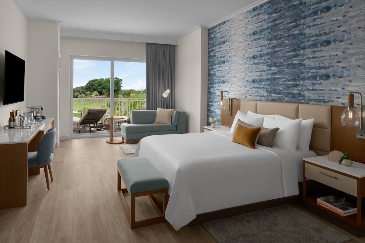 Quarto de hotel com uma cama grande, uma mesa com cadeira, uma mesinha com cadeira e vista para o exterior.