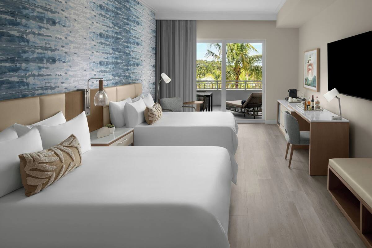 Quarto de hotel com duas camas, uma escrivaninha, uma cadeira, televisão e varanda com vista para palmeiras.