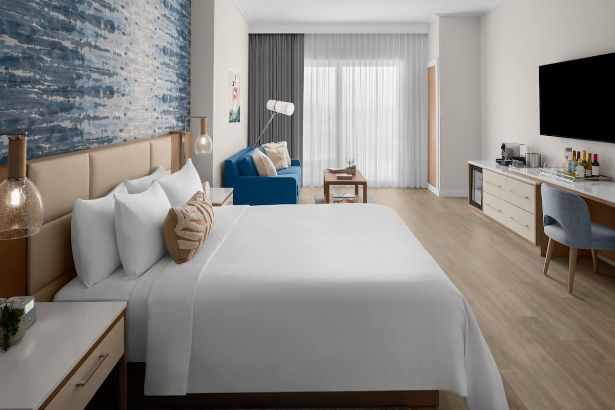 Quarto de hotel moderno com uma cama grande, uma escrivaninha, uma cadeira, uma televisão e uma janela ampla com cortinas.