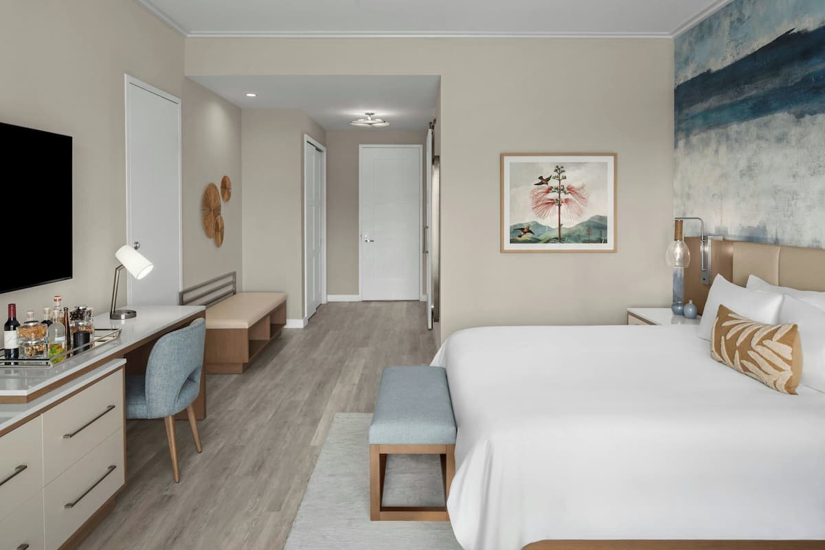 Quarto de hotel com cama, escrivaninha, cadeira, televisão e um quadro na parede.