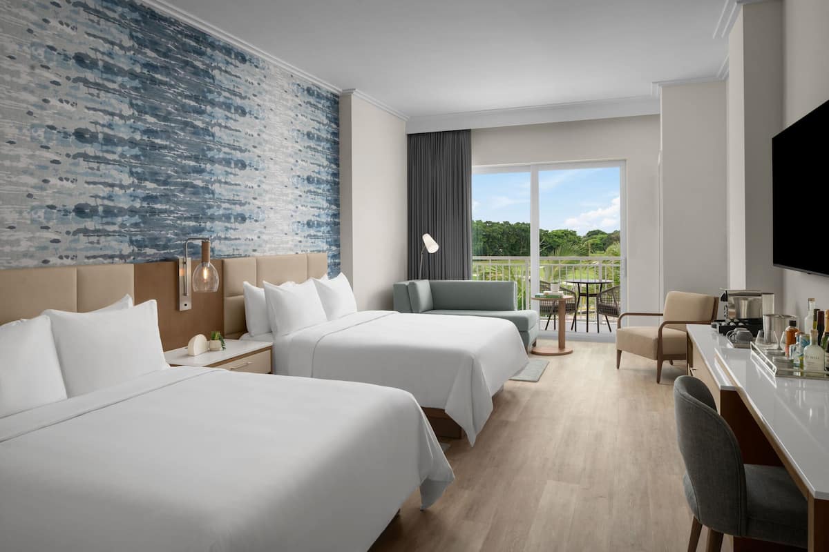 Quarto de hotel com duas camas, área de refeições, televisão e varanda com vista para o verde.