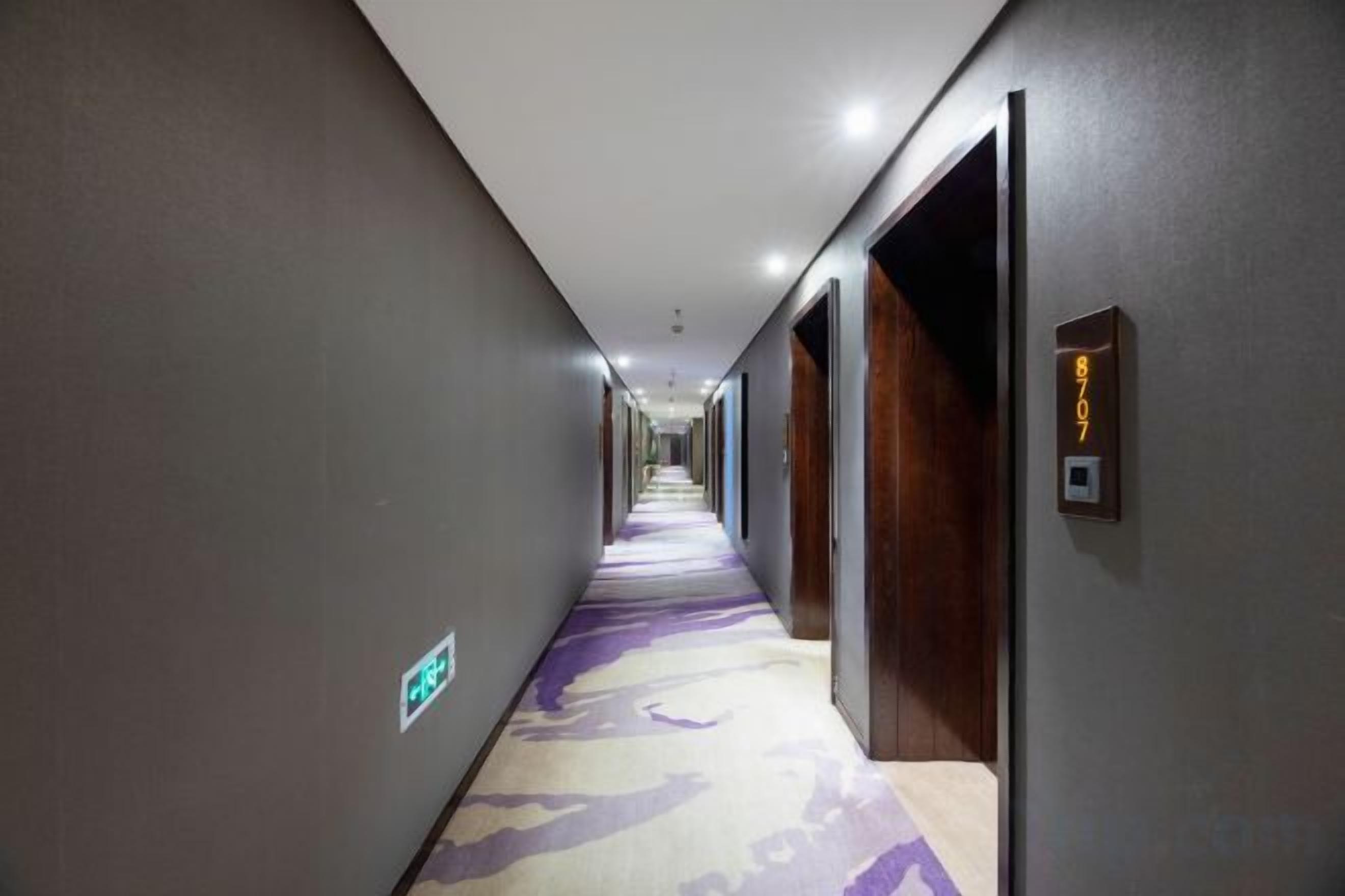Corredor de hotel com piso coberto por carpete, portas à direita e interruptor de luz fixado na parede.