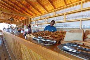 Balcão de pequeno-almoço com diversas opções de comida e bebidas, teto de madeira e prateleiras com copos.