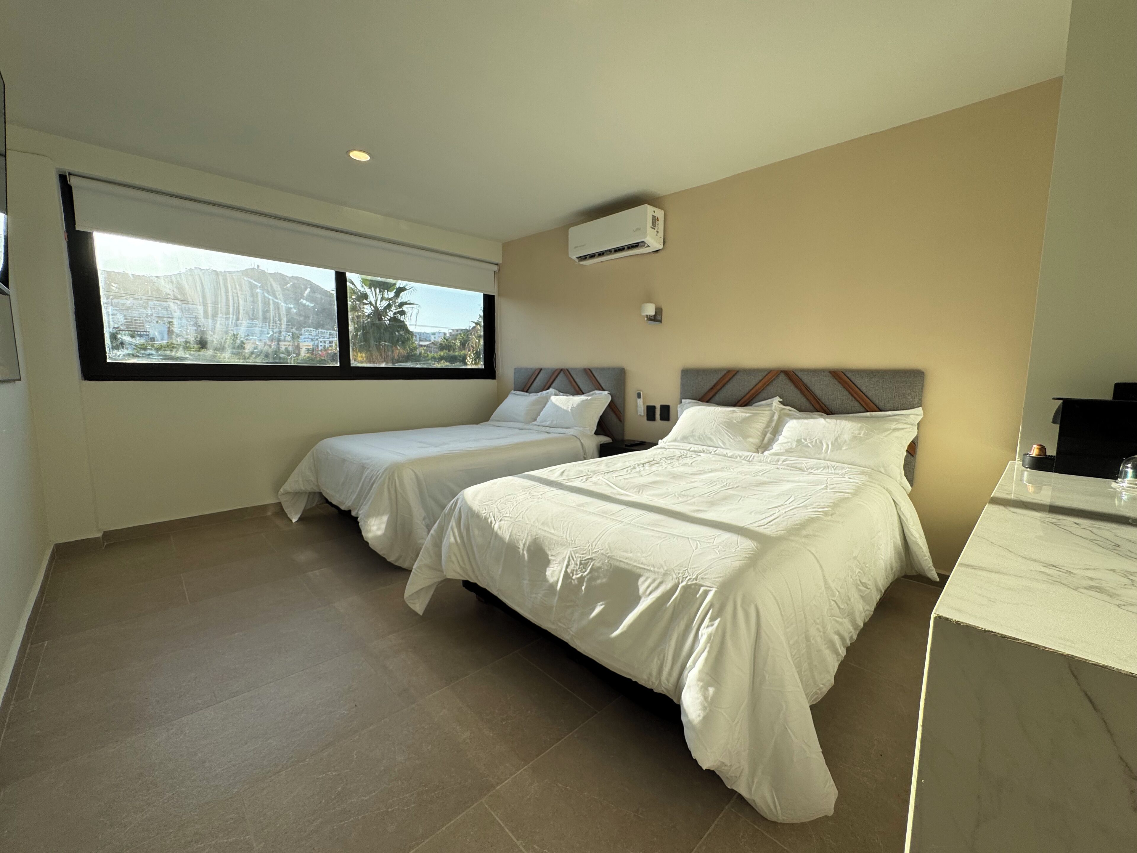 Quarto de hotel com duas camas, uma janela grande, um aparelho de ar condicionado e uma mesa com tampo de mármore.
